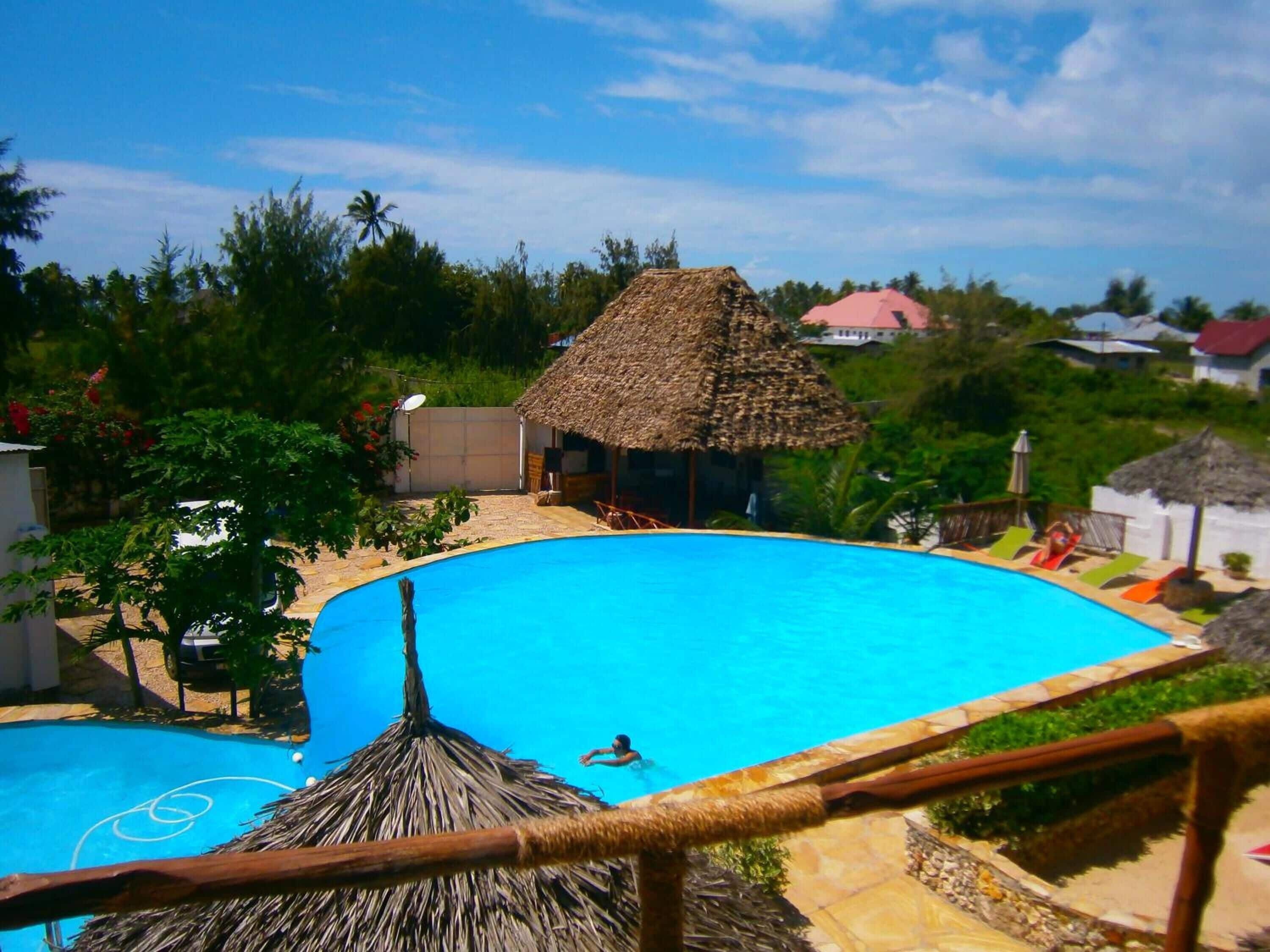Uma piscina com estrutura de telhado de palha, cercada por vegetação e alguns edifícios ao fundo.