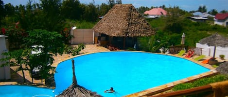Uma piscina com estrutura de telhado de palha, cercada por vegetação e alguns edifícios ao fundo.