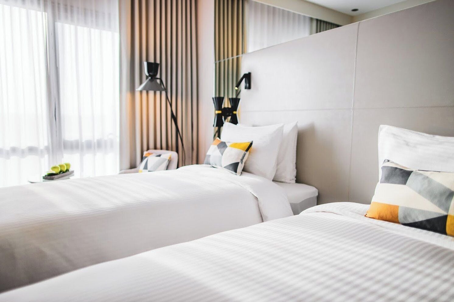 Quarto de hotel com duas camas, uma janela ampla com cortinas leves, luminária de chão e design moderno na parede.