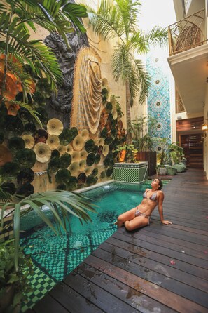 Área de piscina com deck de madeira, cercada por plantas tropicais e elementos decorativos na parede.