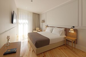 Quarto de hotel moderno com uma cama grande, luminárias laterais, uma TV de tela plana na parede e uma pequena mesa com controle remoto.