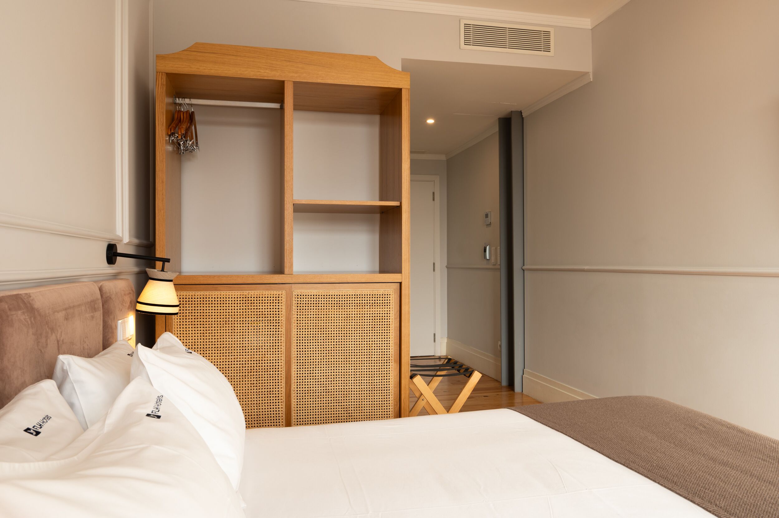 Quarto de hotel com cama, guarda-roupa de madeira, abajur e porta que dá para outro espaço.