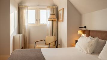 Quarto de hotel com cama, uma cadeira, janela com cortinas, luminária de chão e mesa de cabeceira com abajur.