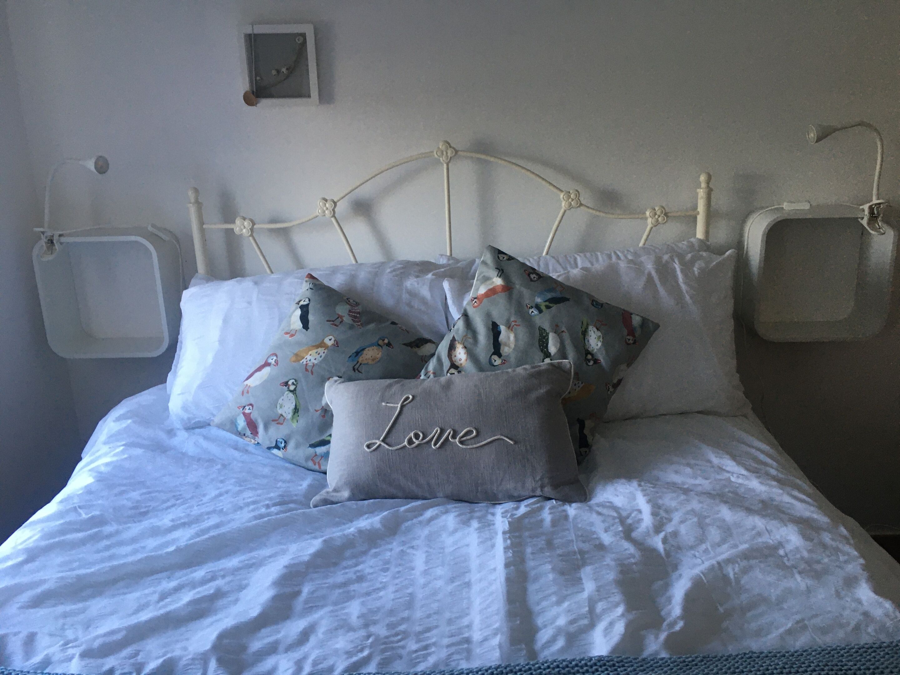 Cama com roupa de cama branca, travesseiro com a inscrição 'Love' e travesseiro com padrão de pássaros.