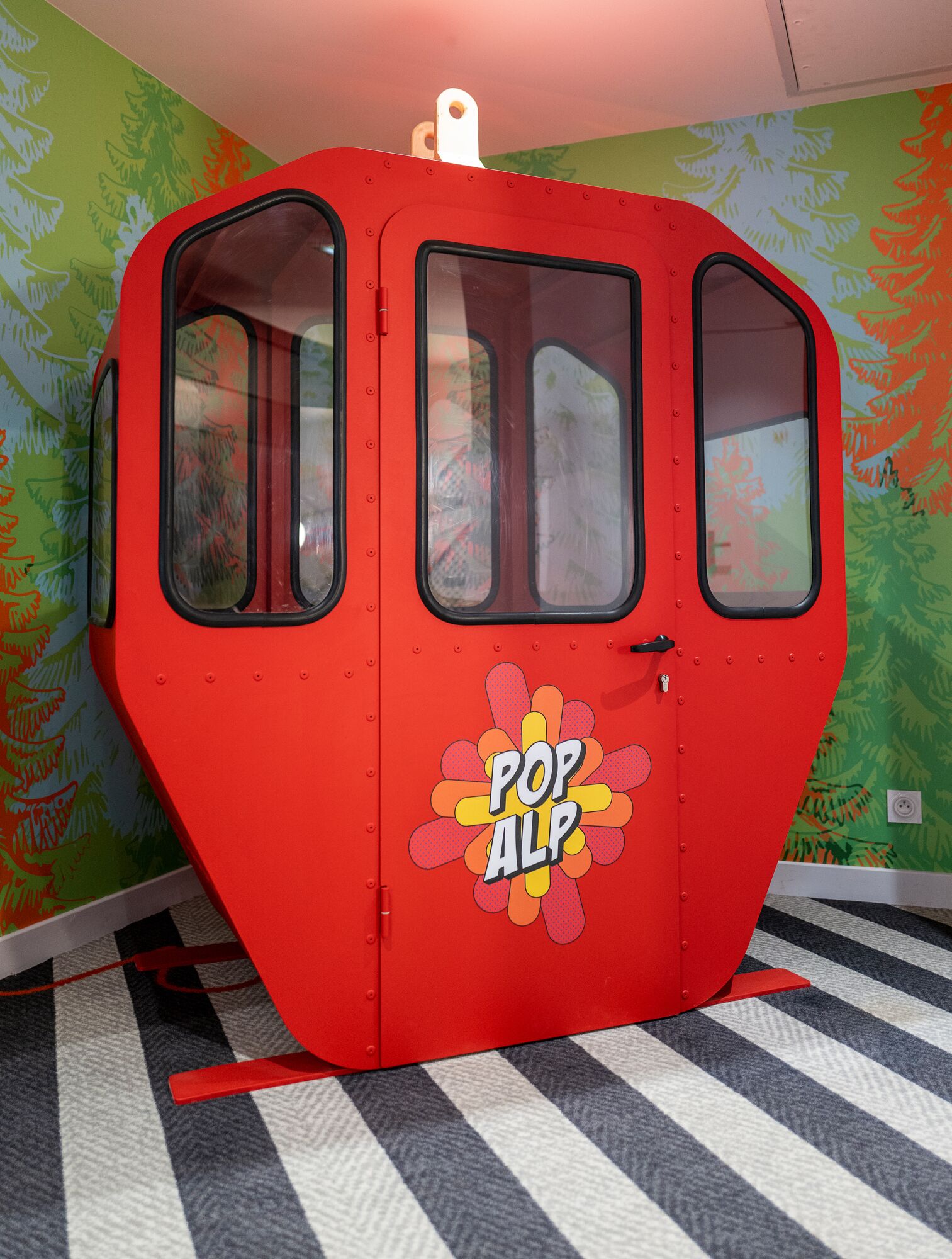 Uma estrutura pop-up vermelha, com janelas e o logotipo 'Pop Alp', montada diante de uma parede colorida com padrões de árvores.