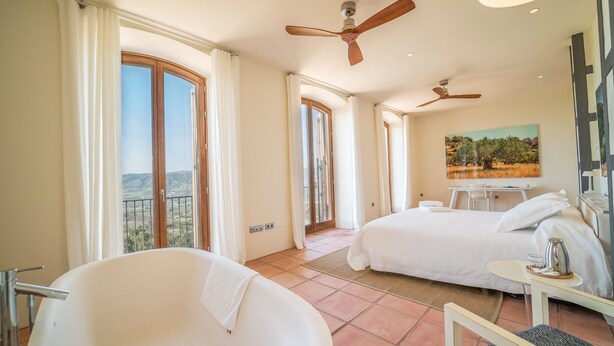 Quarto de hotel com uma cama grande, área de estar com uma poltrona, escrivaninha e uma pintura de paisagem. Há duas portas que dão para varandas com vista para o exterior.
