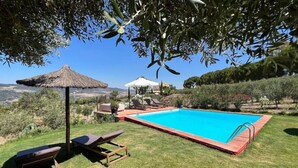 Piscina com espreguiçadeiras e um guarda-sol de palha, cercada por vegetação e vista aberta para a paisagem.