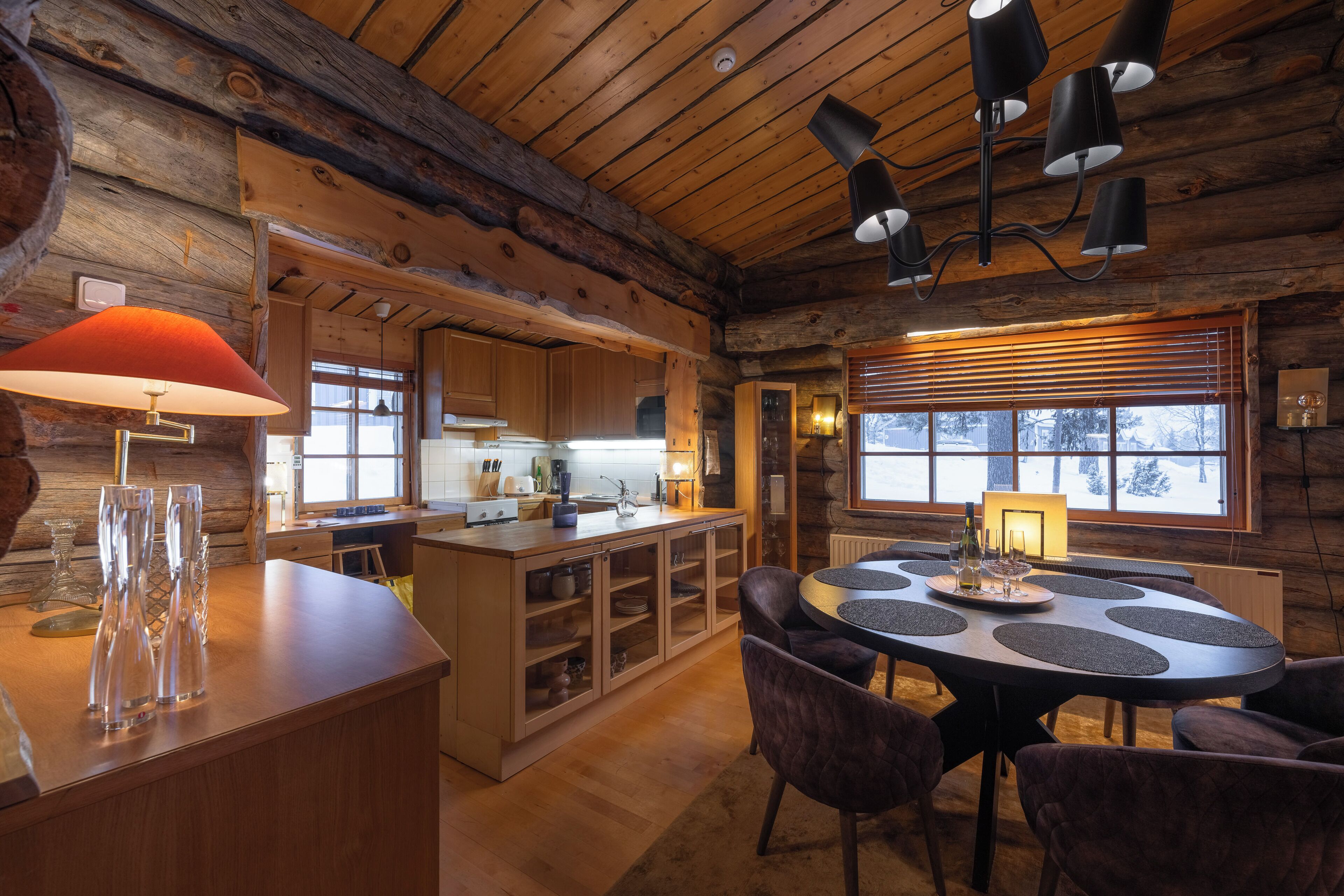 Interior de uma cabana de troncos, com uma mesa de jantar de madeira, cadeiras, área de cozinha e uma grande janela com vista para árvores.