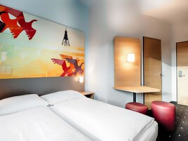 Quarto de hotel com cama, um banquinho vermelho, luz acoplada à parede e uma grande pintura abstrata.