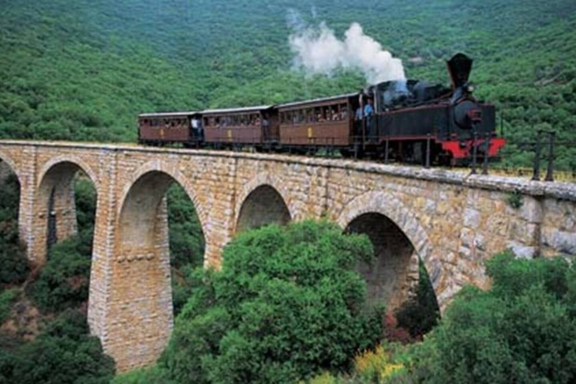 Uma locomotiva a vapor atravessando uma ponte de pedra com várias arcadas, rodeada por vegetação exuberante.
