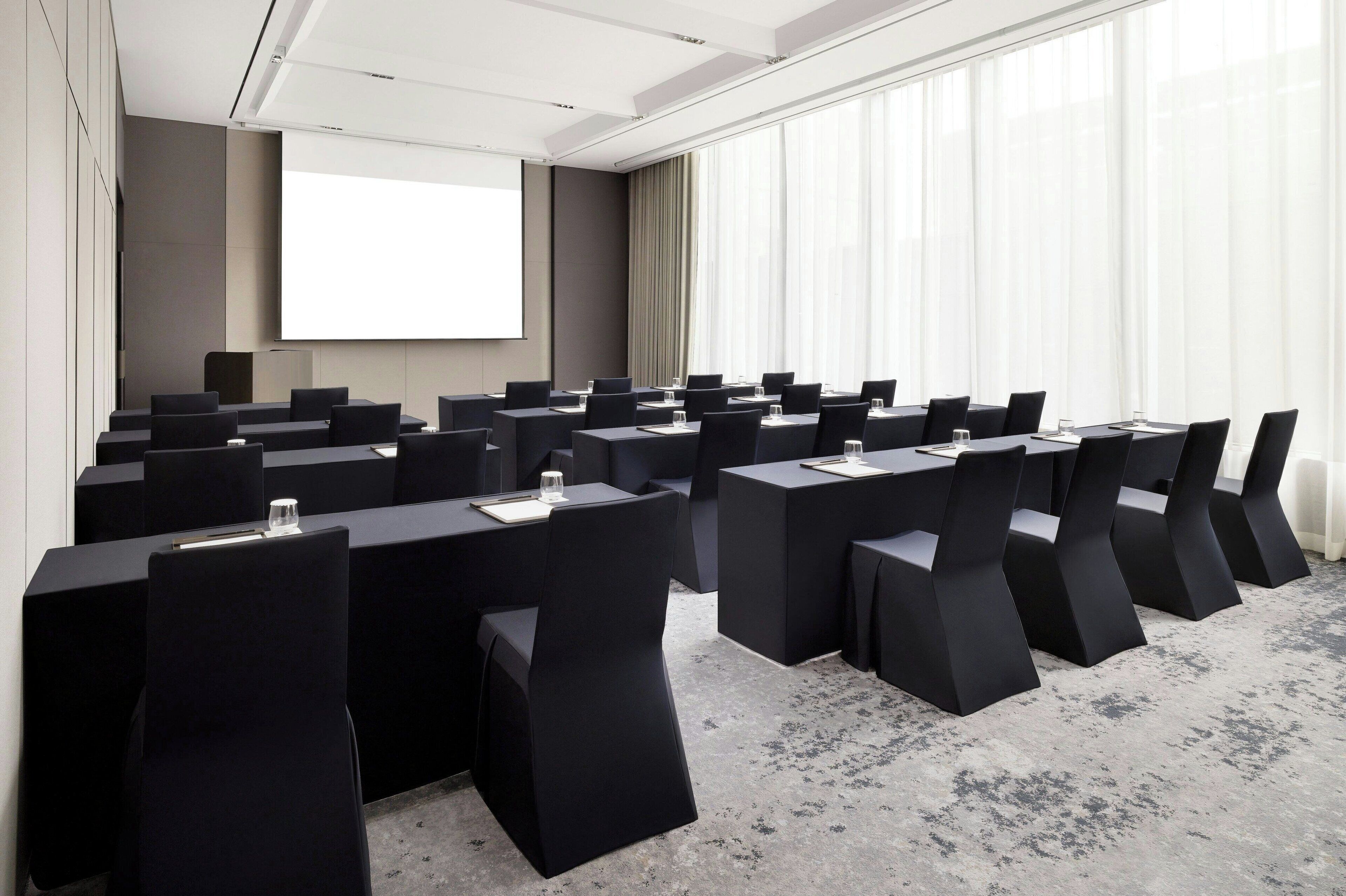 Sala de conferências com várias mesas e cadeiras, uma tela de projetor e janelas amplas com cortinas leves.