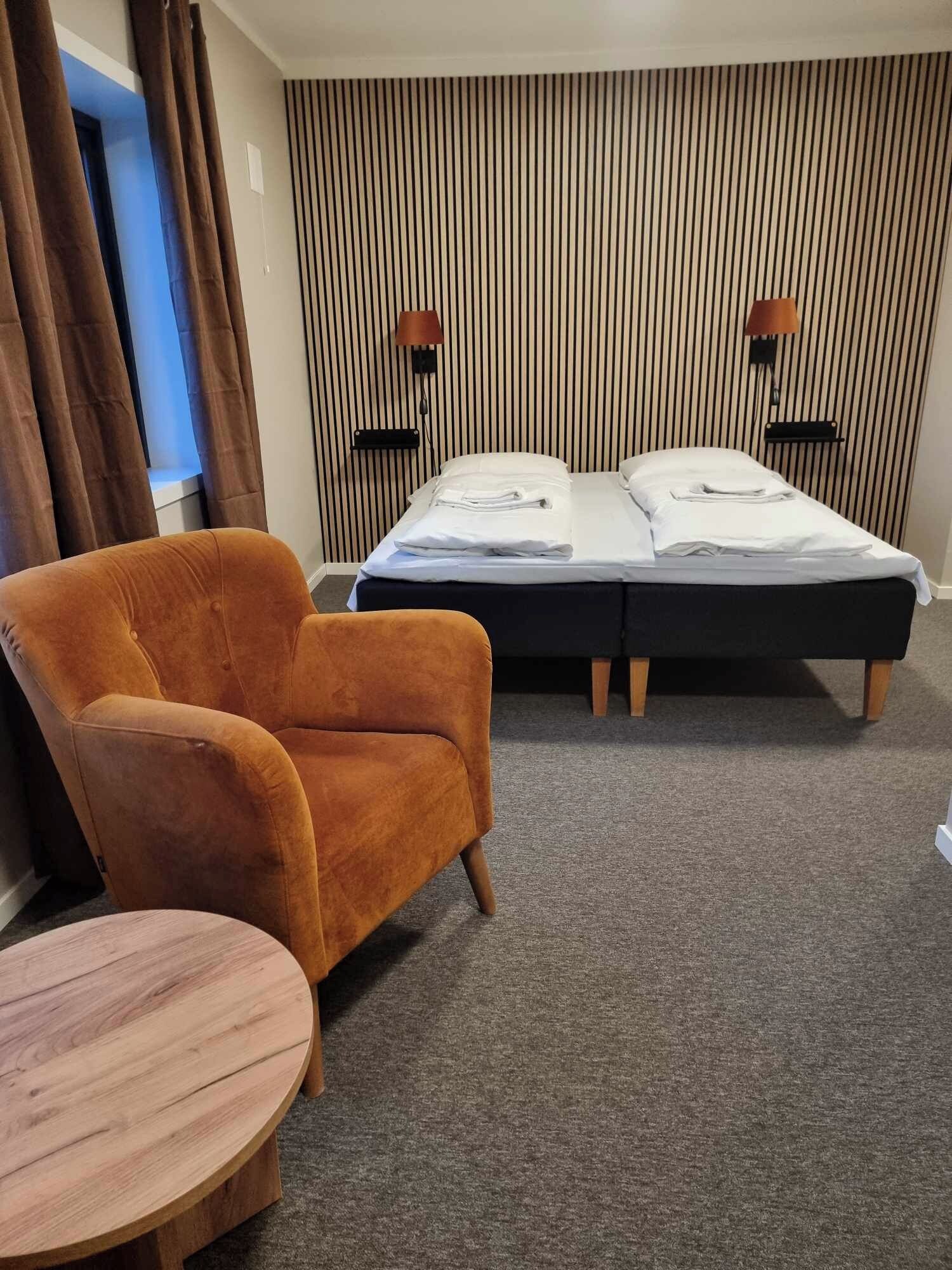 Quarto de hotel com uma cama de solteiro, uma cadeira e uma mesinha.