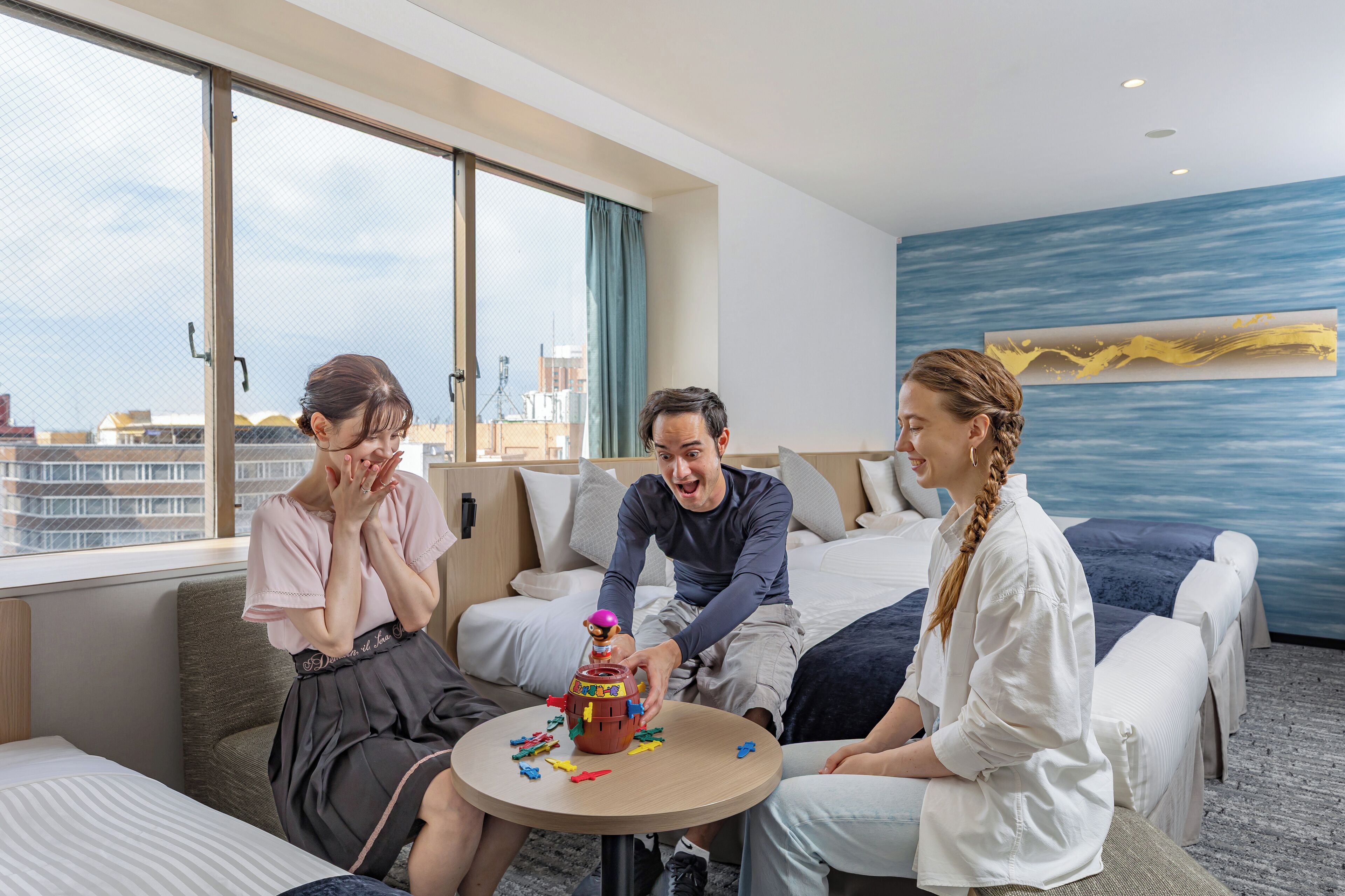 Quarto de hotel com uma janela grande, uma mesa redonda com brinquedos e uma cama com roupa de cama branca.