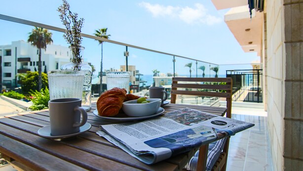 Uma mesa de madeira com jornal, um croissant, uma tigela de azeitonas e um copo de água numa varanda com vista para a cidade.