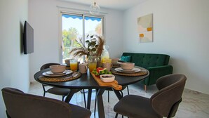 Uma sala de jantar com uma mesa posta para quatro pessoas, rodeada por cadeiras. Um sofá verde está encostado à parede, e um quadro está pendurado acima.