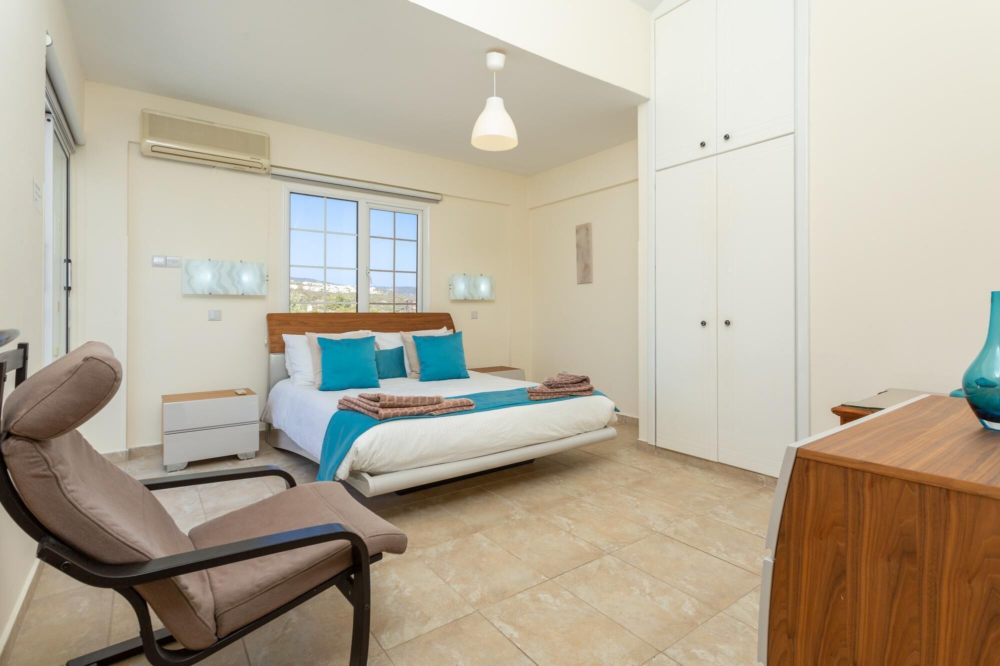 Um quarto com cama, mesas de cabeceira, uma cadeira, escrivaninha e guarda-roupa.