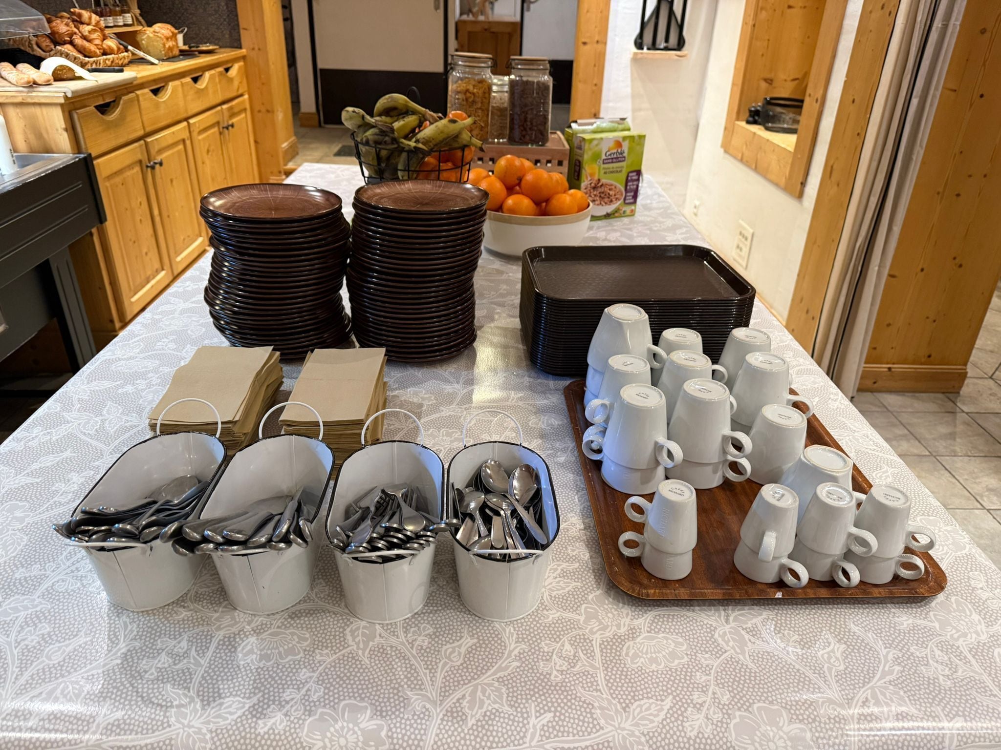 Uma bancada de cozinha com pilhas de pratos, tigelas e canecas, juntamente com uma bandeja de laranjas e caixas de cereais.