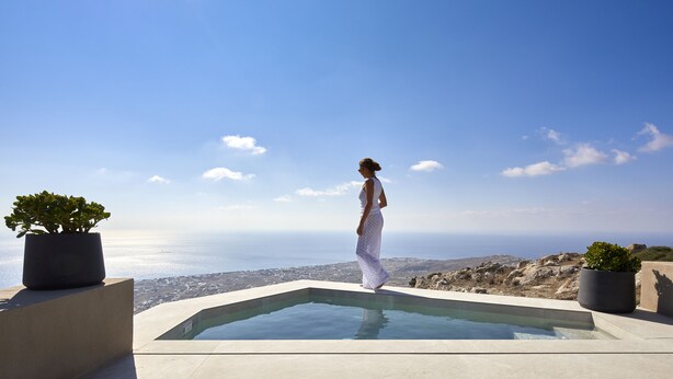 Uma pessoa parada junto a uma piscina com vista para o mar e paisagem distante.