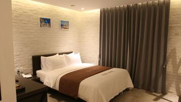 Quarto de hotel com parede de tijolos brancos, uma cama com roupa de cama branca e um cobertor marrom, uma mesa de cabeceira com um telefone e uma janela com cortinas.