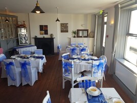 Uma sala de jantar com mesas preparadas para uma refeição, cadeiras com fitas azuis, lareira e janela com cortinas.