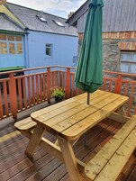Um deck de madeira com mesa de piquenique e um guarda-sol verde, cercado por uma cerca de madeira e um muro de pedra.