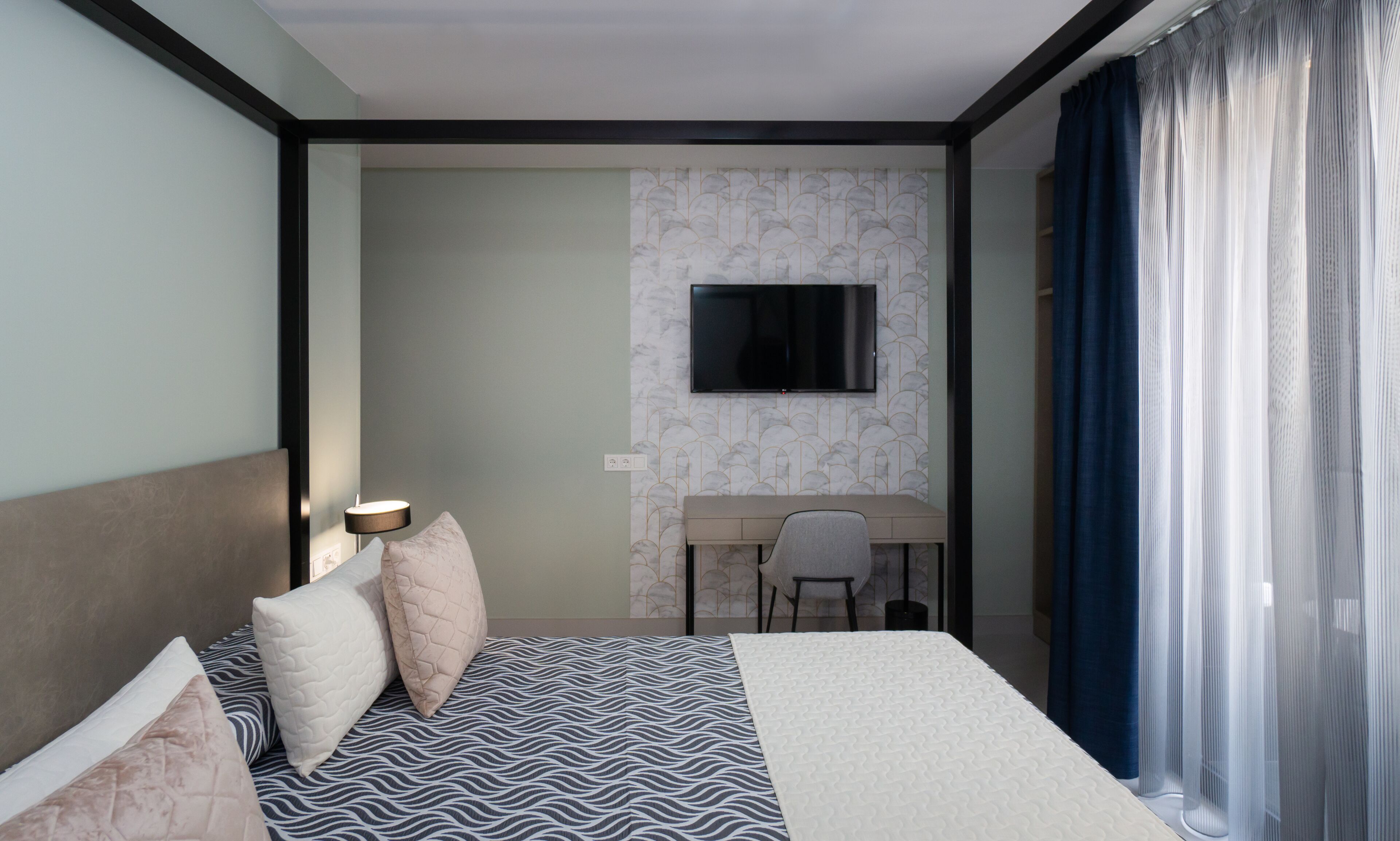 Quarto de hotel com uma cama, uma escrivaninha, uma cadeira e uma televisão na parede.