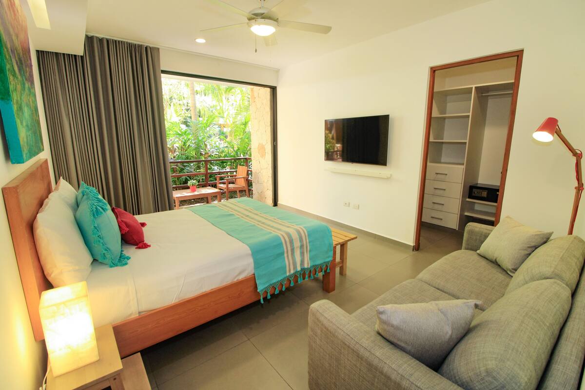 Quarto com cama, sofá, televisão, ventoinha no teto e porta de correr para uma varanda.