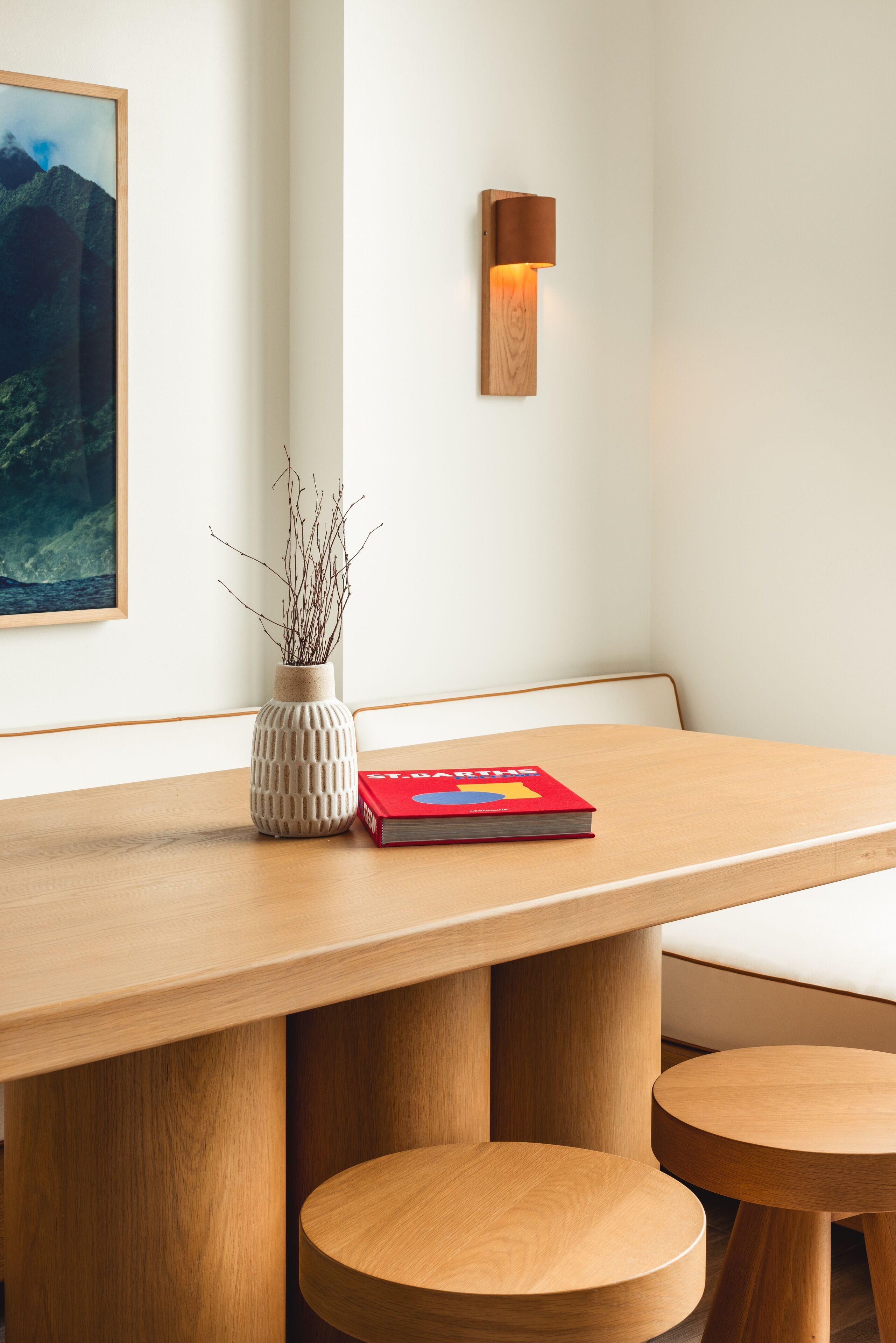 Uma mesa de madeira com um vaso e um livro, uma luminária na parede e um quadro enquadrado.