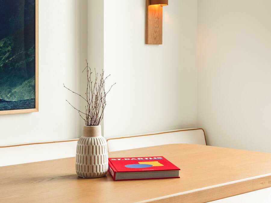 Uma mesa de madeira com um vaso e um livro, uma luminária na parede e um quadro enquadrado.