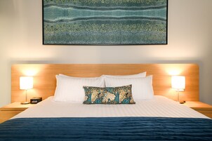 Cama bem arrumada com colcha azul, almofadas brancas e uma almofada decorativa com padrão inspirado na natureza. Duas luminárias de cabeceira em ambos os lados da cama, e uma obra de arte emoldurada acima.
