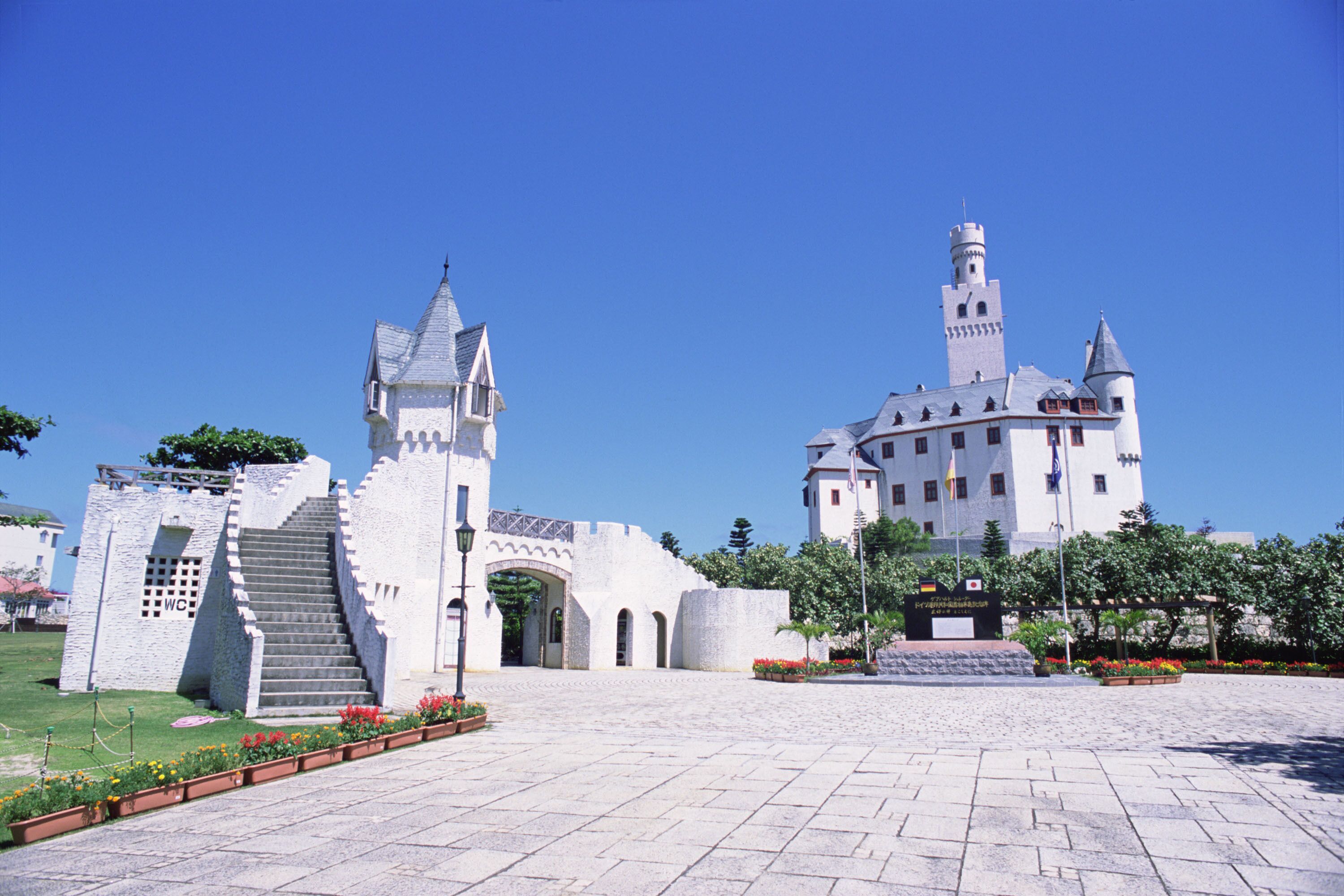 Um edifício semelhante a um castelo, com uma torre alta e outra menor, rodeado por um pátio pavimentado com canteiros de flores.