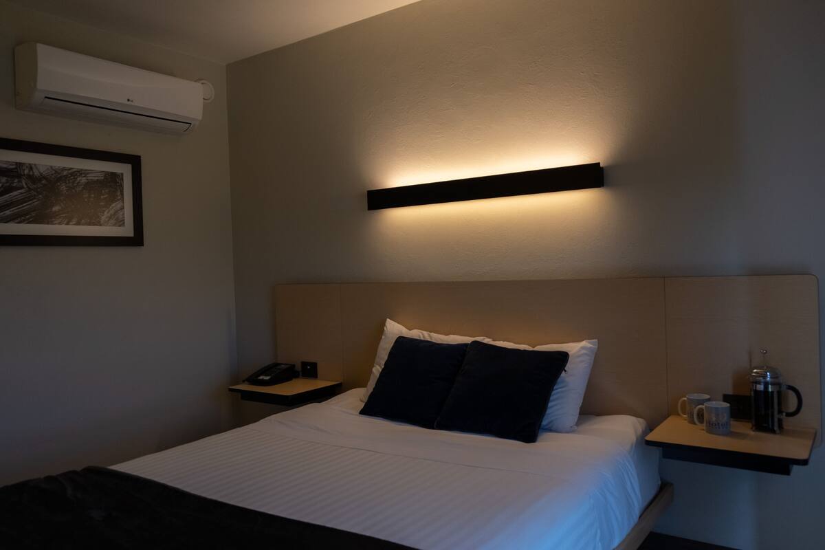 Quarto de hotel com cama, mesas de cabeceira, ar condicionado fixado na parede, um quadro emoldurado e abajur.