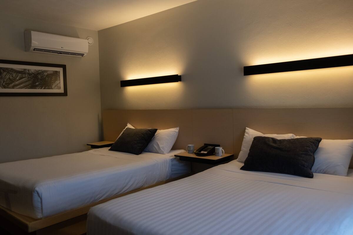 Quarto de hotel com duas camas, mesa de cabeceira com telefone e caneca, ar condicionado e um quadro na parede.