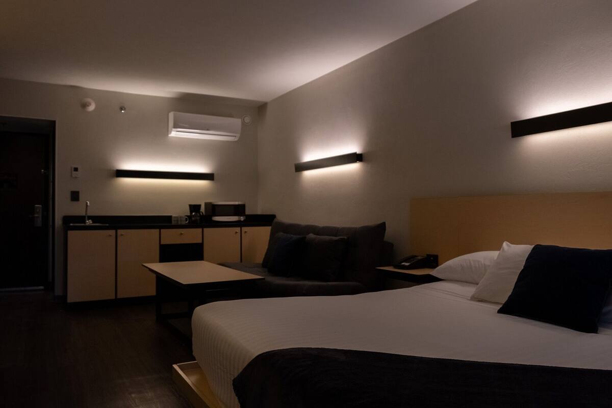 Quarto de hotel com cama, escrivaninha, cozinha compacta e ar condicionado fixado na parede.
