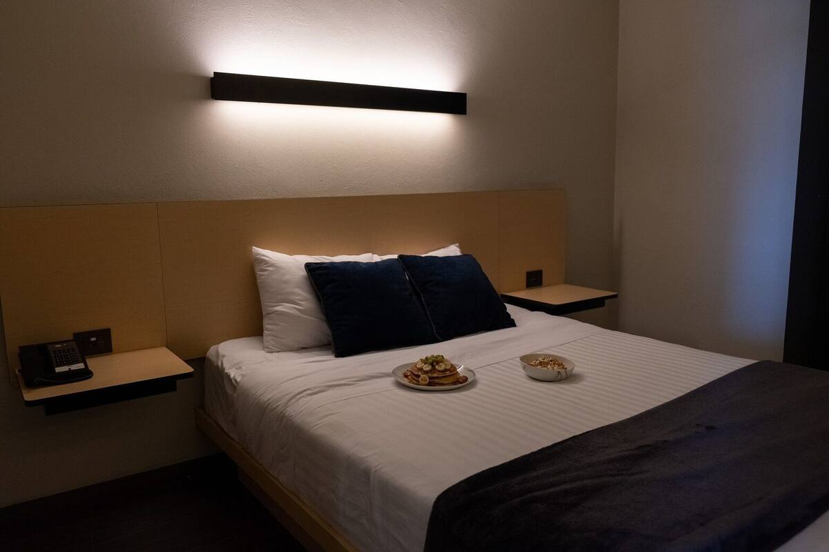 Quarto de hotel com cama, duas mesas de cabeceira, luminária na parede e uma pequena mesa com telefone e bloco de notas.