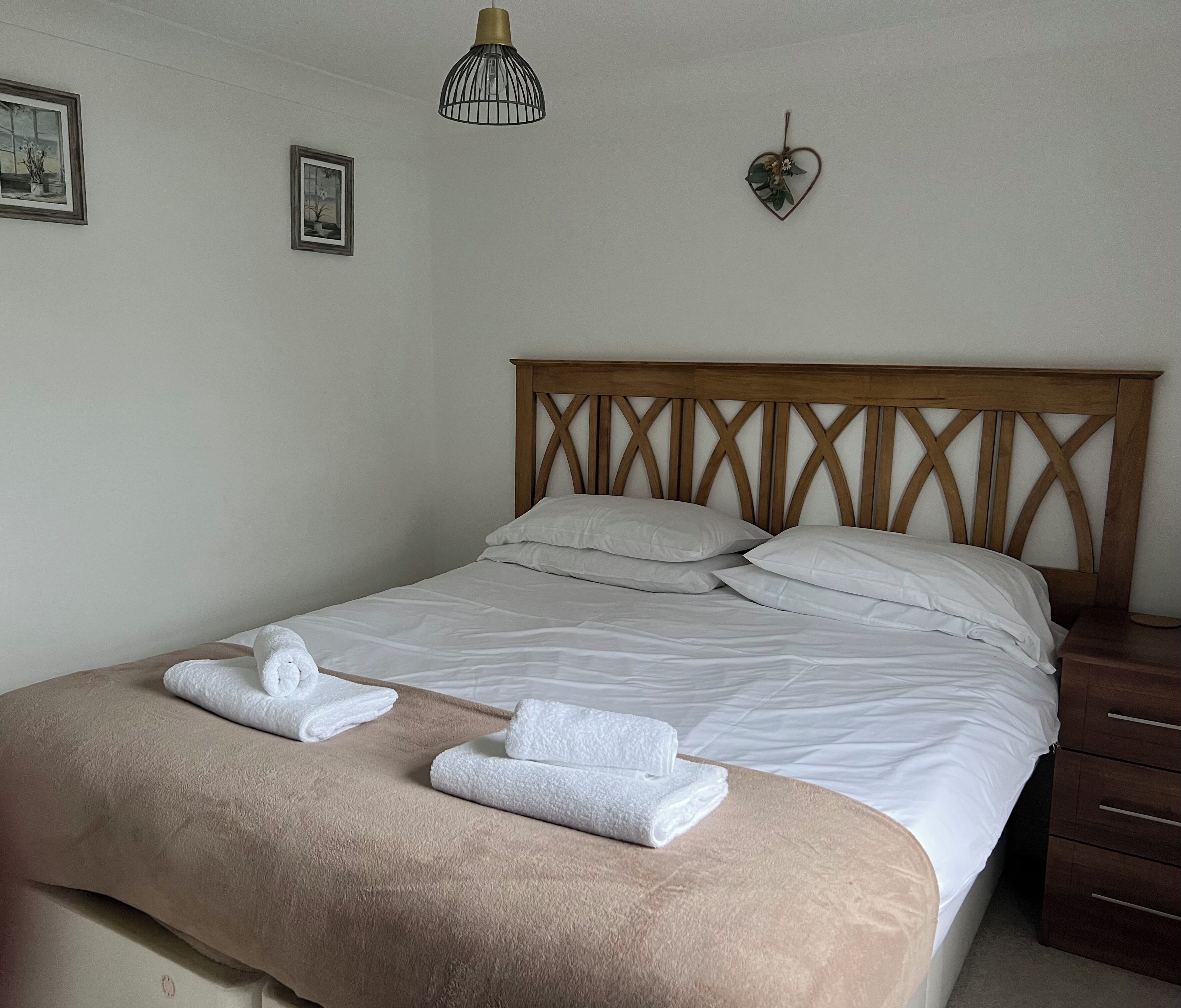 Cama bem arrumada com lençóis brancos e manta marrom, cabeceira de madeira e luminária pendente.