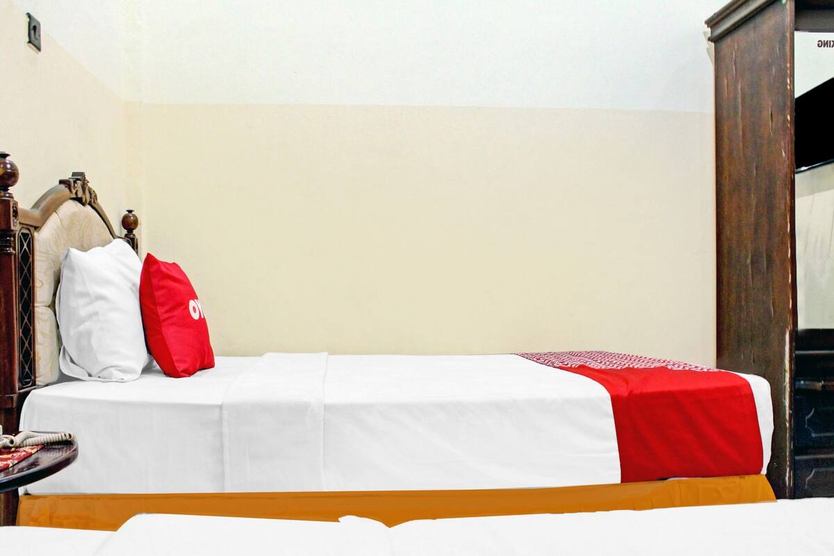 Uma cama de solteiro com colcha branca e vermelha, cabeceira de madeira e uma mesinha de cabeceira com telefone e um objeto vermelho ao lado.