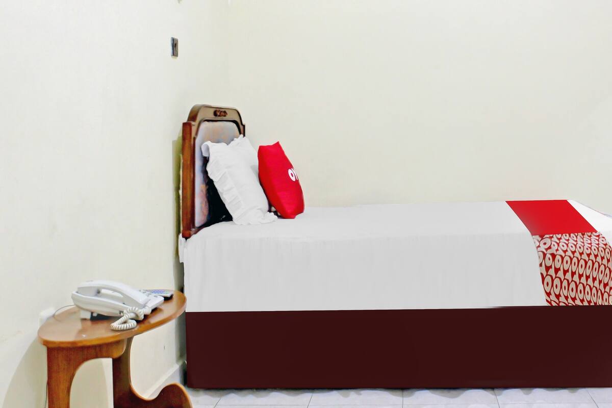 Uma cama de solteiro com um cobreleiro branco e vermelho, um painel de cabeceira em madeira, um travesseiro vermelho e um telefone de mesa branco sobre um criado-mudo de madeira.