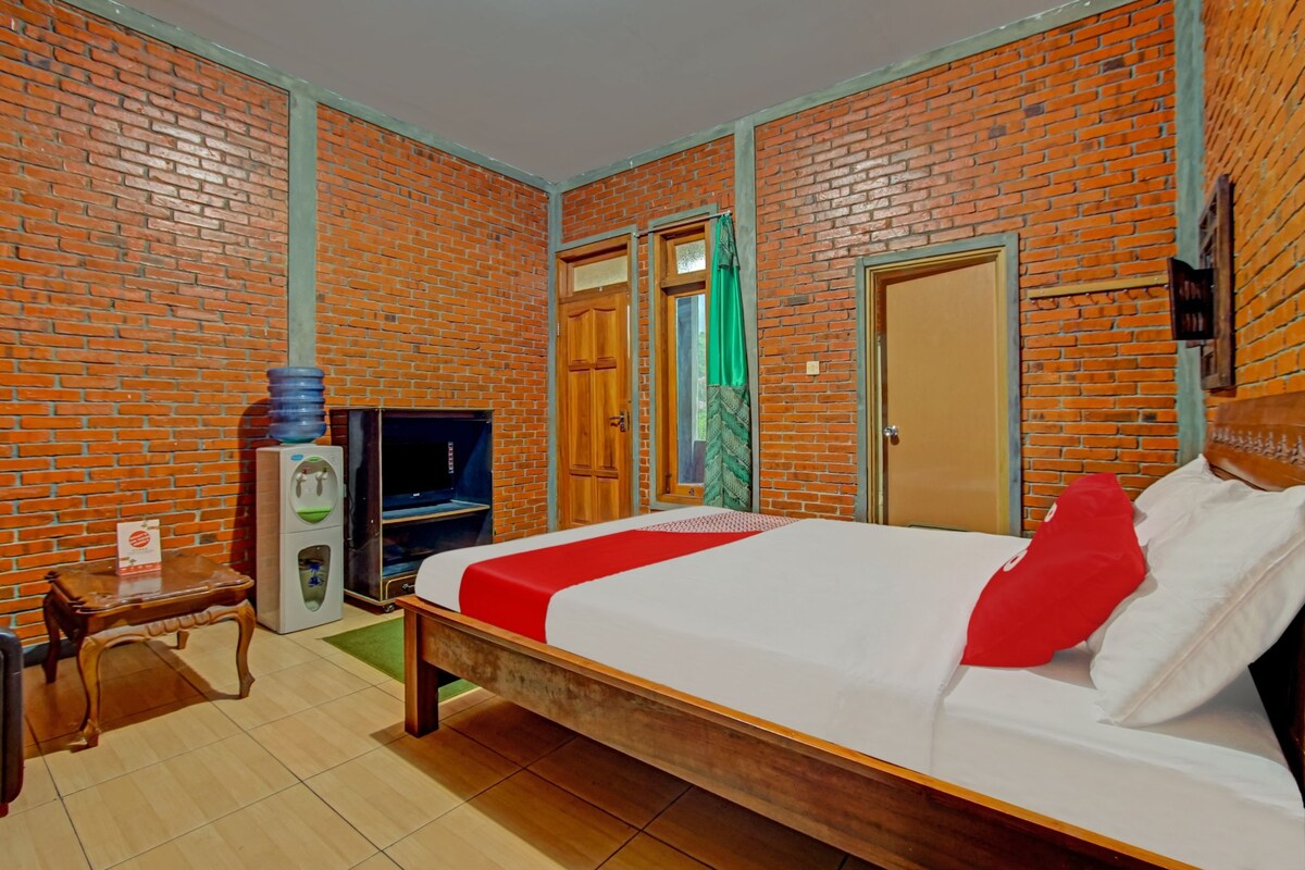 Quarto com parede de tijolos, cama com roupa de cama branca e vermelha, um banco de madeira, uma mesa pequena e uma televisão.