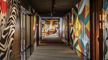 Um corredor de hotel com paredes em padrão de zebra, piso coberto por carpete e um grande mural de um rosto na parede ao fundo.