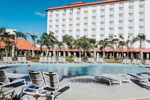 Hotel com piscina, espreguiçadeiras, palmeiras e um edifício grande com sacadas.
