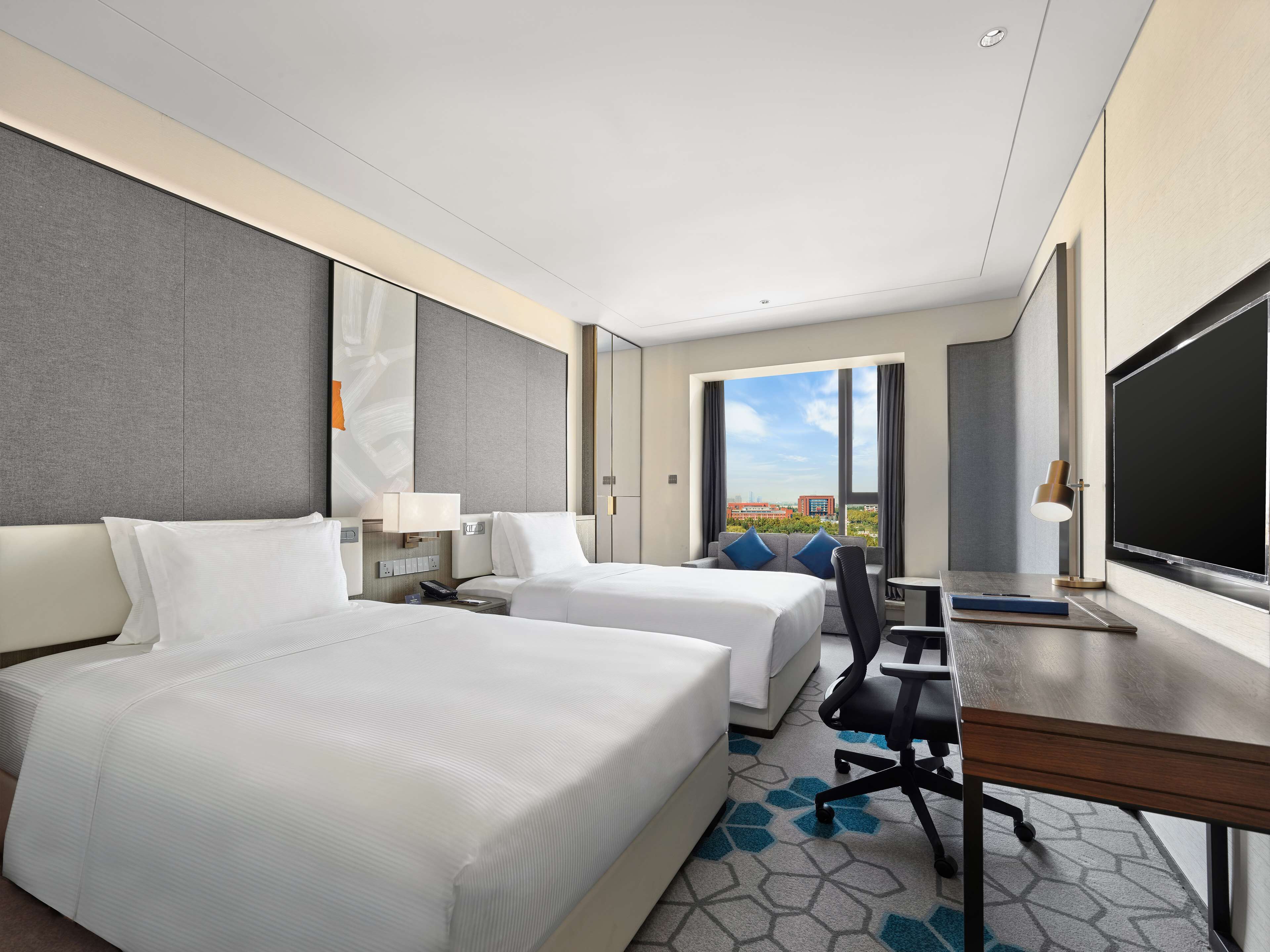 Quarto de hotel com duas camas, uma escrivaninha, uma cadeira, uma televisão e uma janela com vista para a cidade.