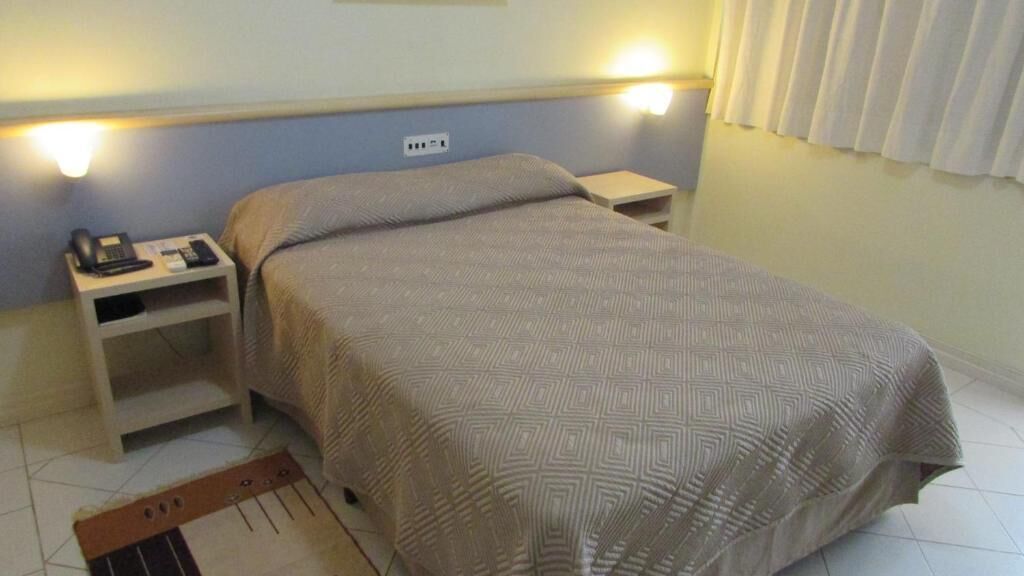 Uma cama de solteiro com colcha estampada, mesas de cabeceira com abajures e uma tomada na parede.