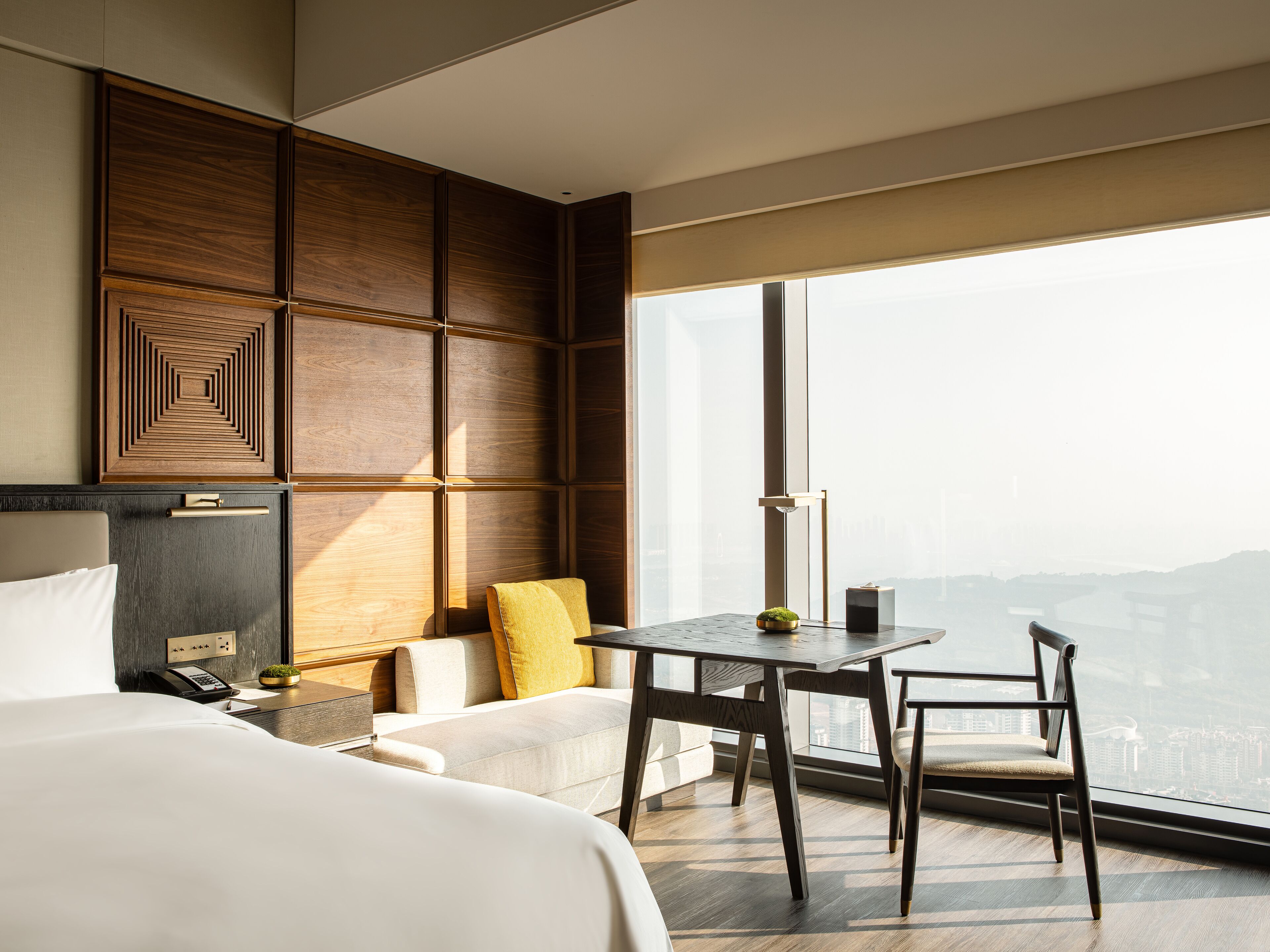 Quarto de hotel moderno com cama grande, área de estar, mesa de jantar e vista para a cidade.