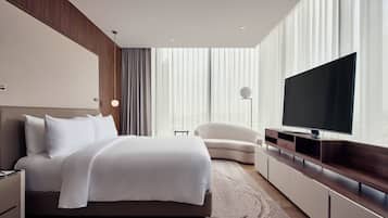 Quarto de hotel moderno com uma cama grande, televisão de tela plana, sofá e uma janela ampla com cortinas.