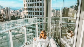 Uma pessoa sentada num sofá branco com guarda-corrimão de vidro, com vista para um horizonte urbano com edifícios altos.