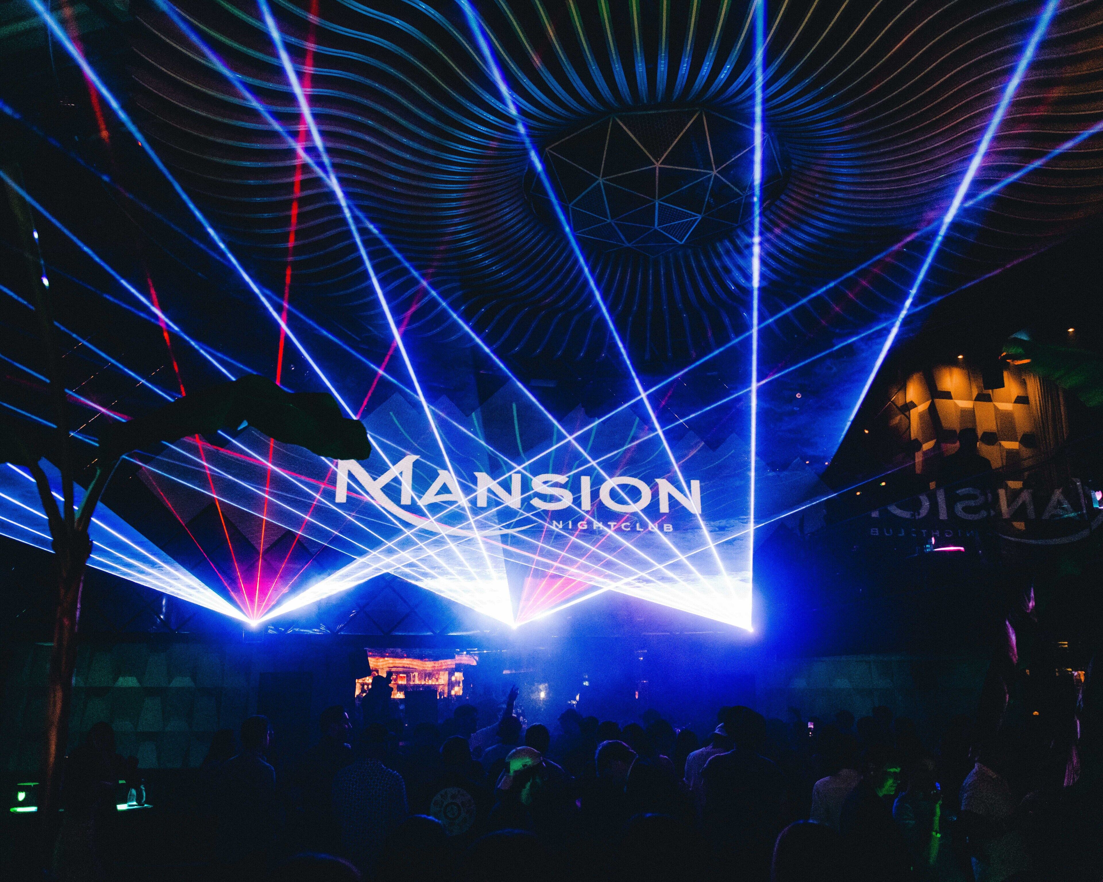 Interior de uma discoteca com um letreiro chamativo 'Mansion', padrões de luz geométricos e uma multidão de pessoas.