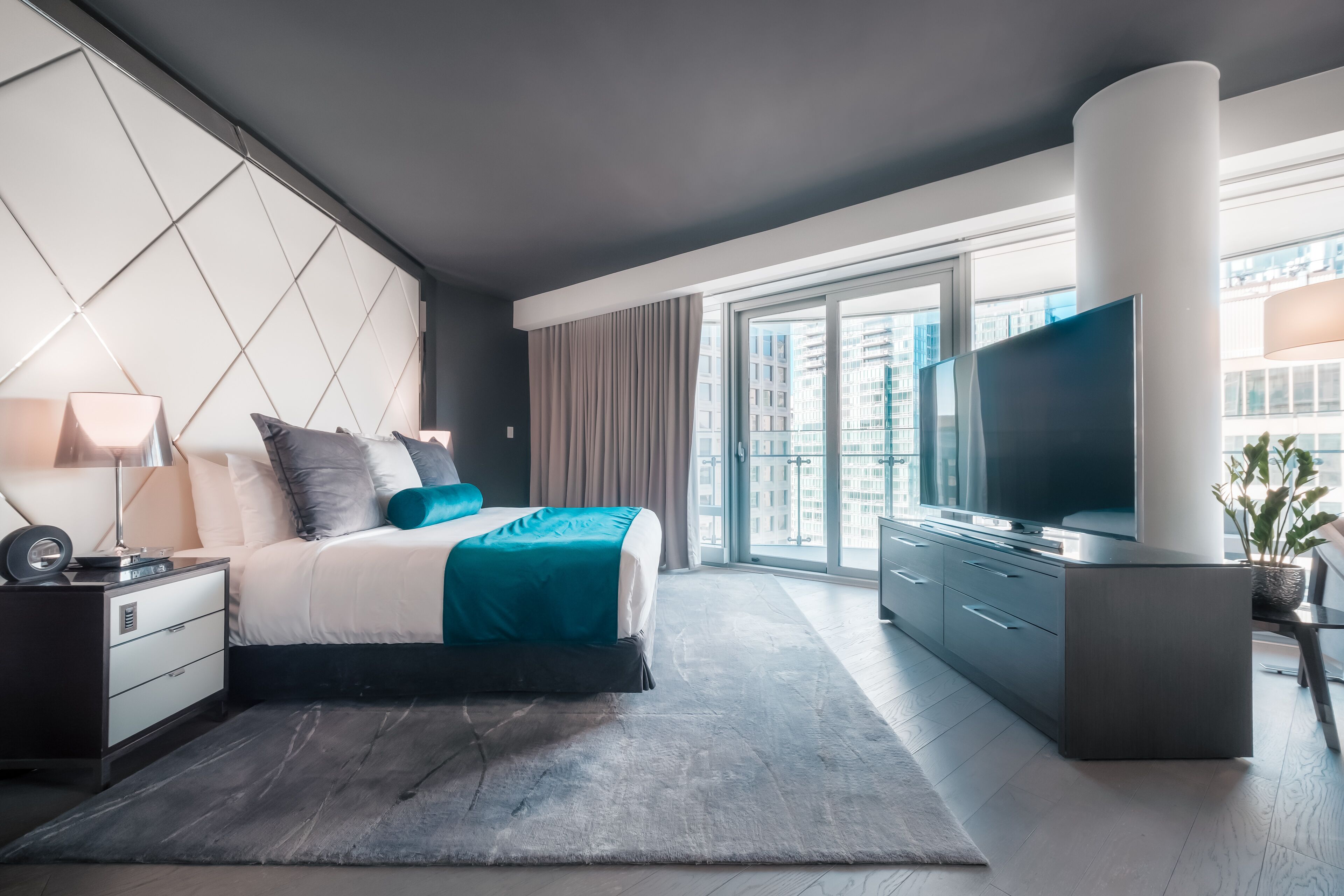 Quarto de hotel moderno com uma cama grande, uma TV de tela plana, uma mesa de cabeceira e vista para a cidade através de amplas janelas.