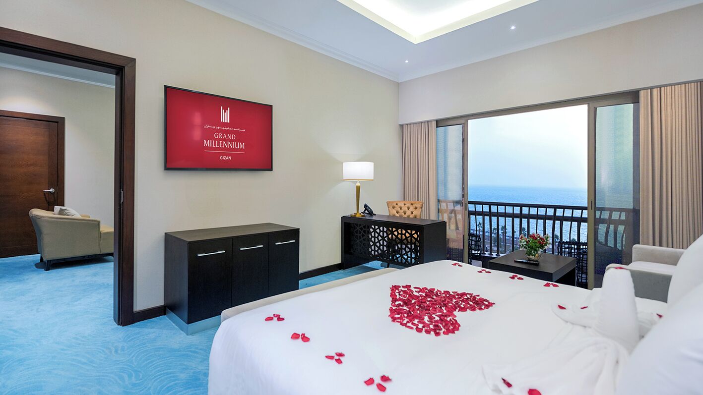 Quarto de hotel com cama, televisão, escrivaninha, cadeira e varanda com vista para o mar.