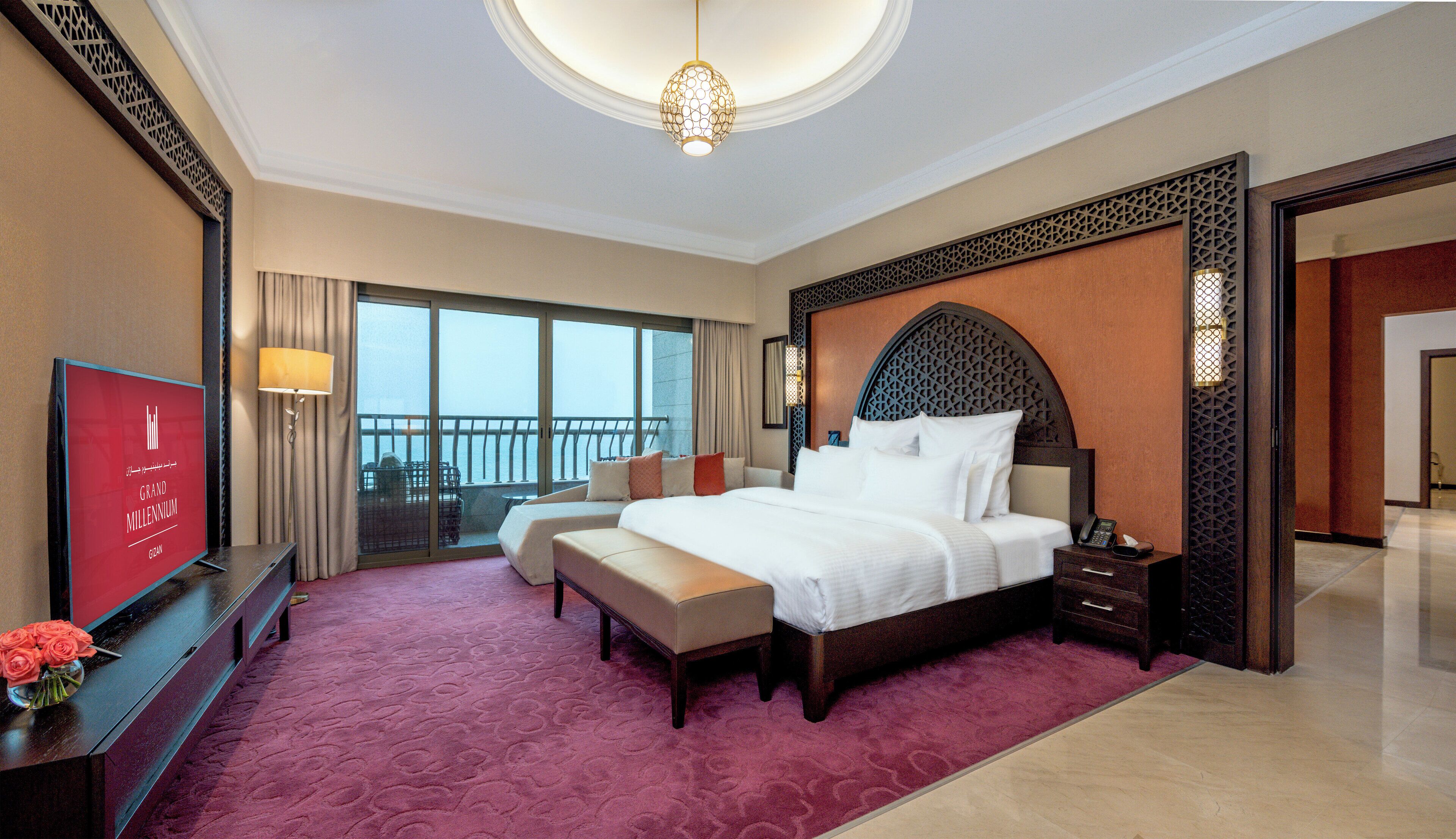 Quarto de hotel com uma cama grande, uma escrivaninha com televisão, varanda com vista e cabeceira decorativa.